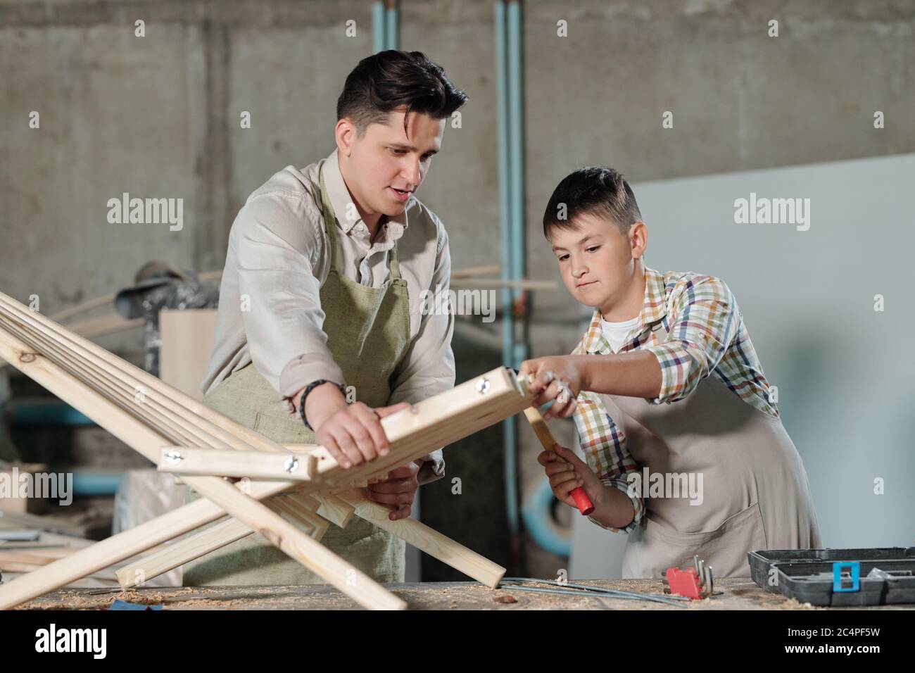 Skilled teenage boy in apron using hammer while working on wooden ...