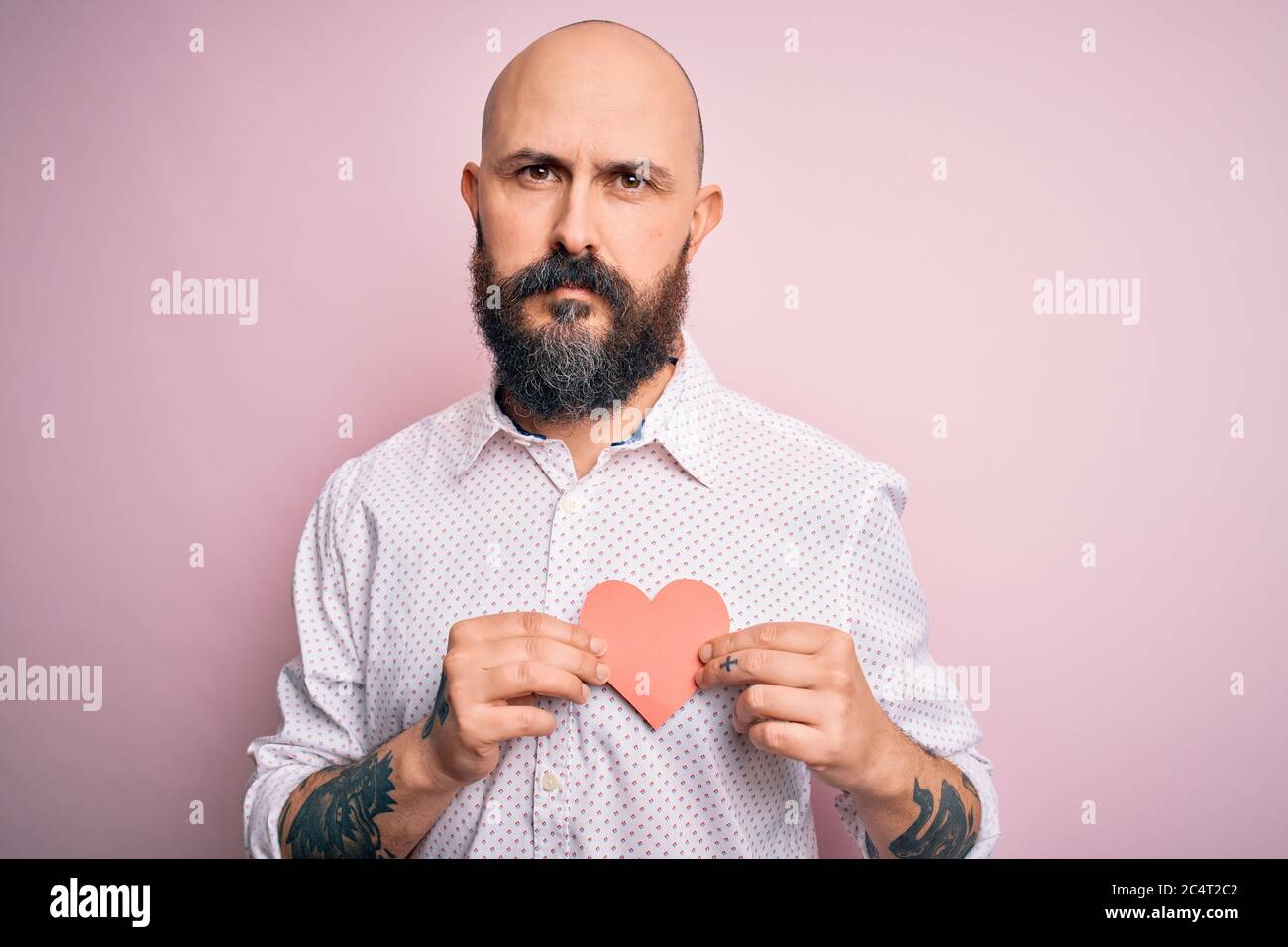 Handsome romantic bald man with beard holding red heart paper over pink ...