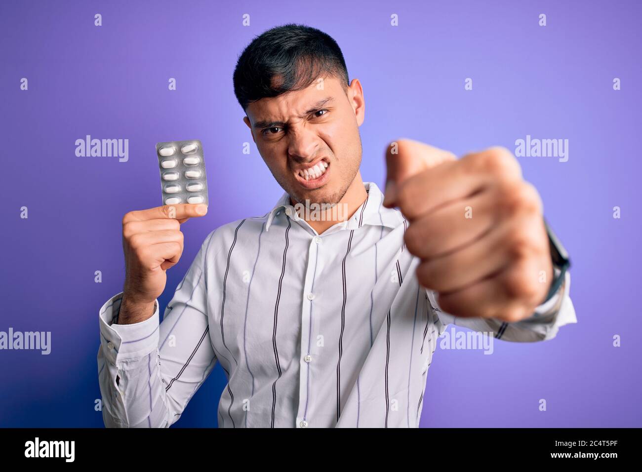 Young hispanic man holding pharmaceutical antibiotics pills over purple ...