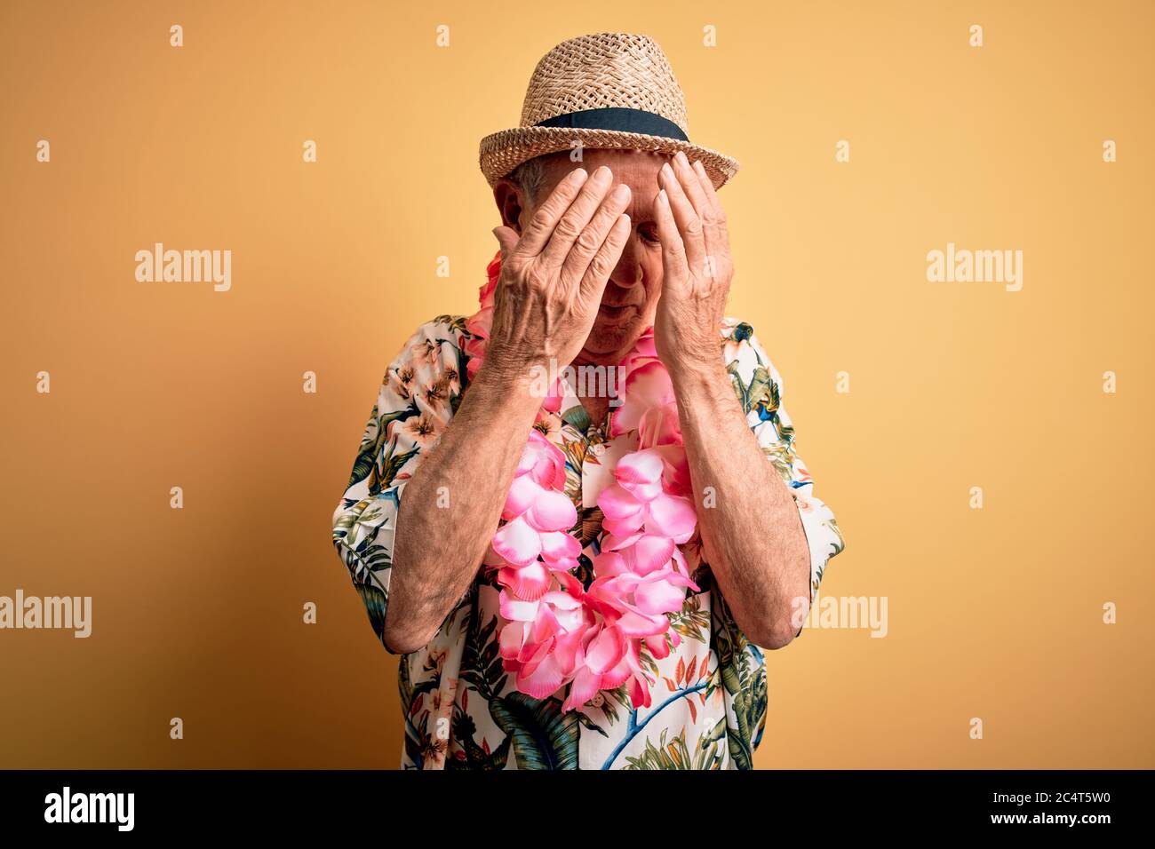 Grey haired senior man wearing summer hat and hawaiian lei over yellow ...