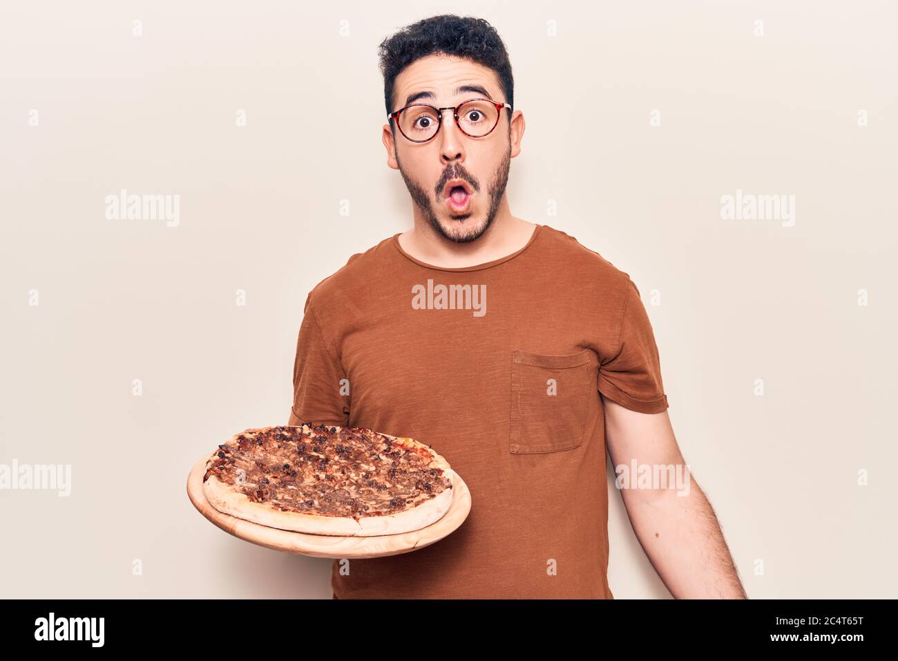 Young hispanic man wearing glasses holding italian pizza scared and ...