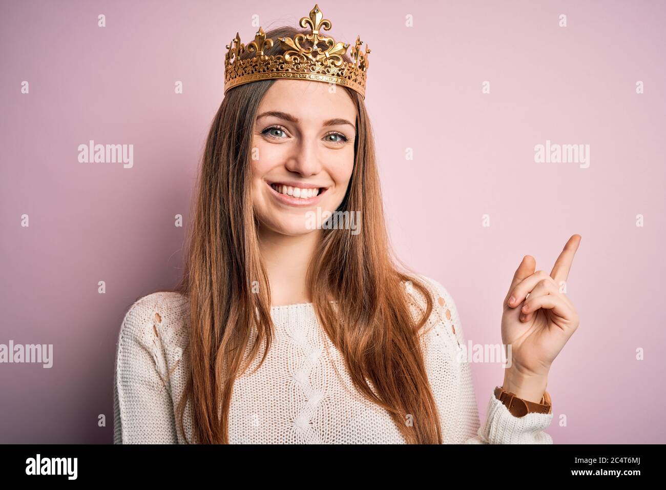 Young beautiful redhead woman wearing queen crown over isolated pink ...