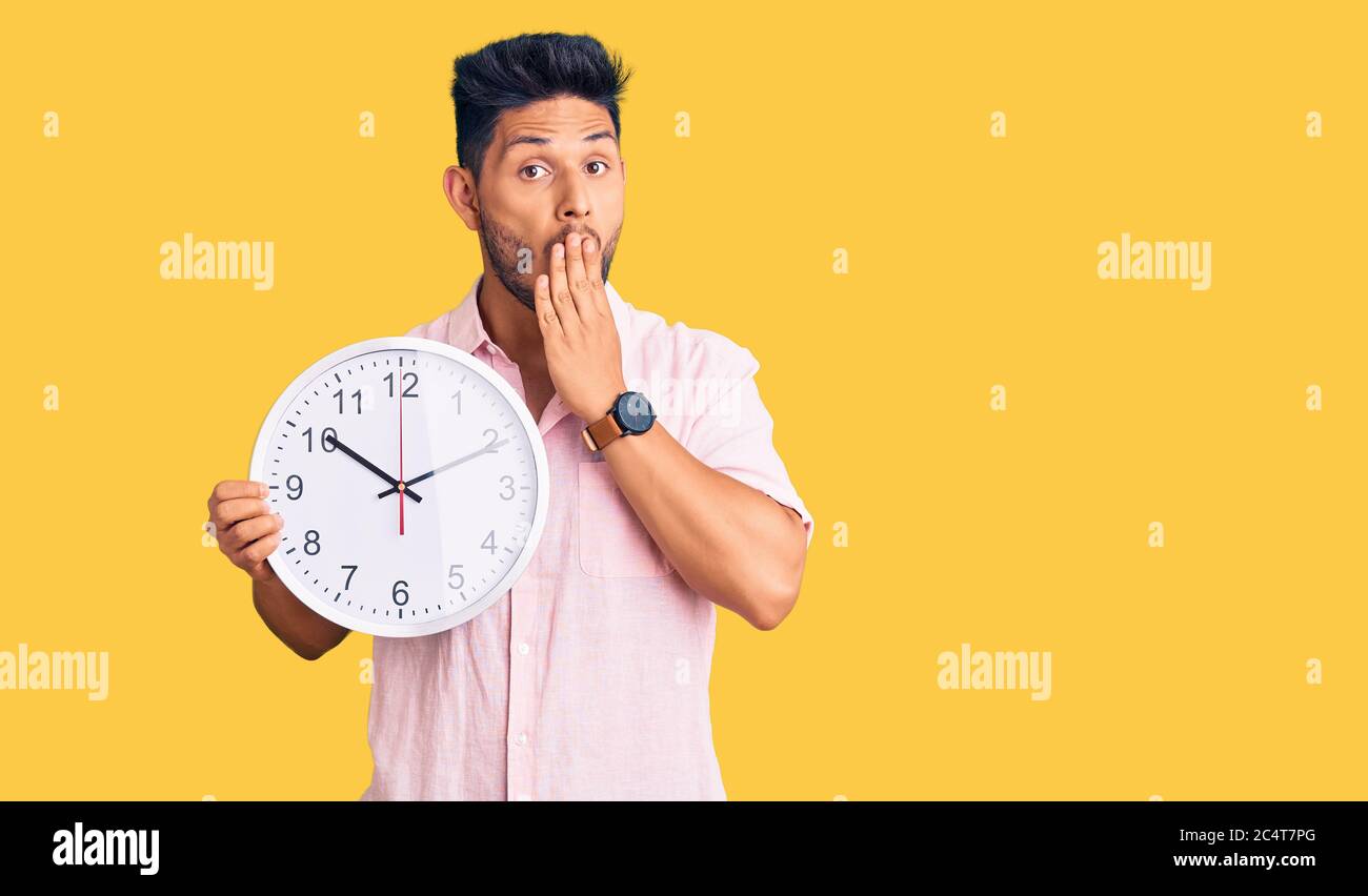 Handsome latin american young man holding big clock covering mouth with ...