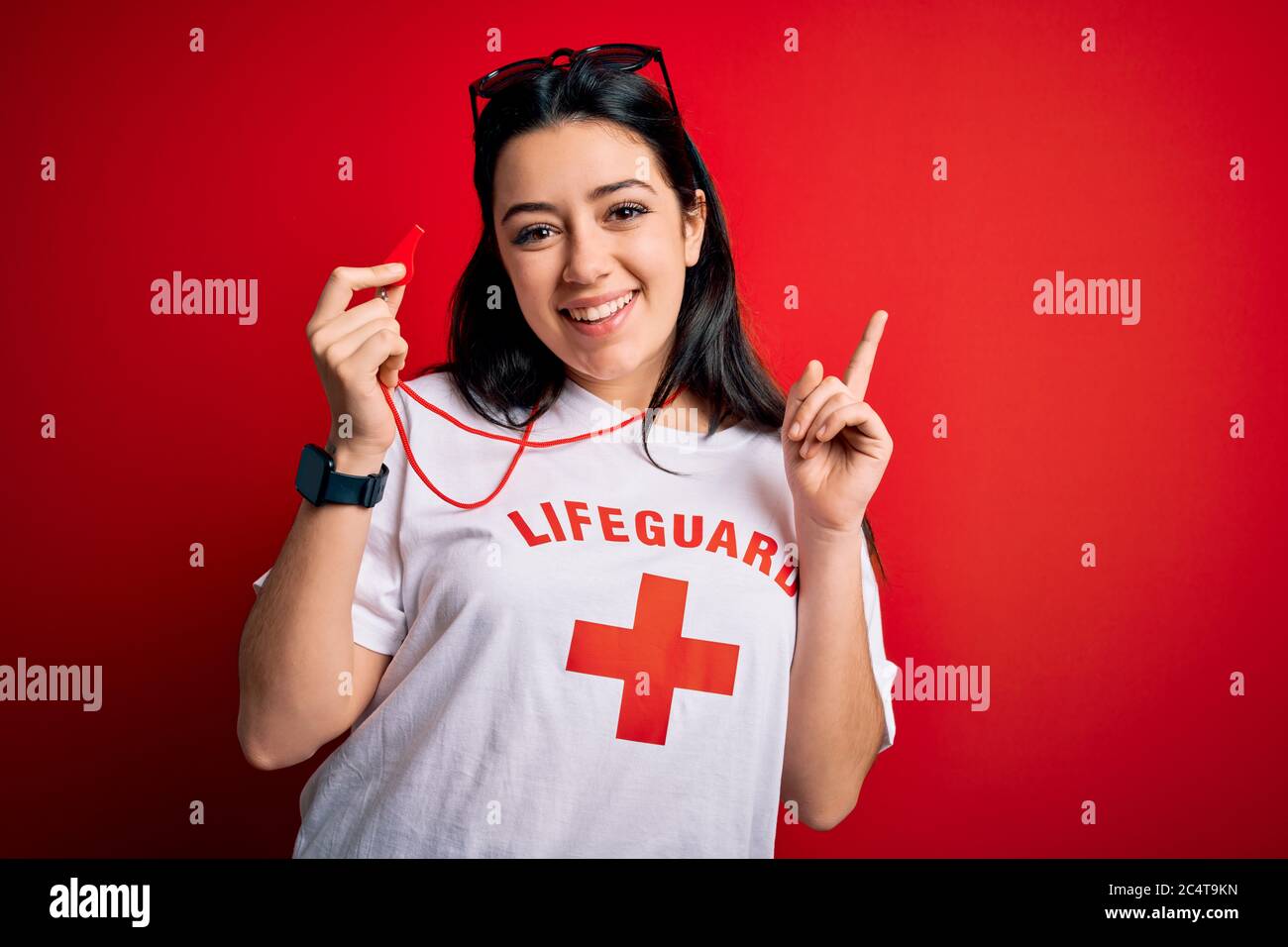 Young lifeguard woman wearing guard equipement holding whistle over red ...