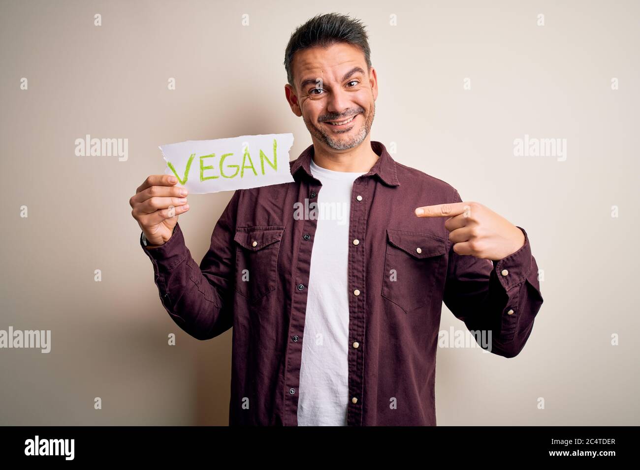 Young handsome veggie man holding paper with vegan message over white ...