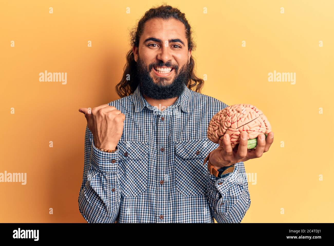 Young arab man holding brain pointing thumb up to the side smiling ...
