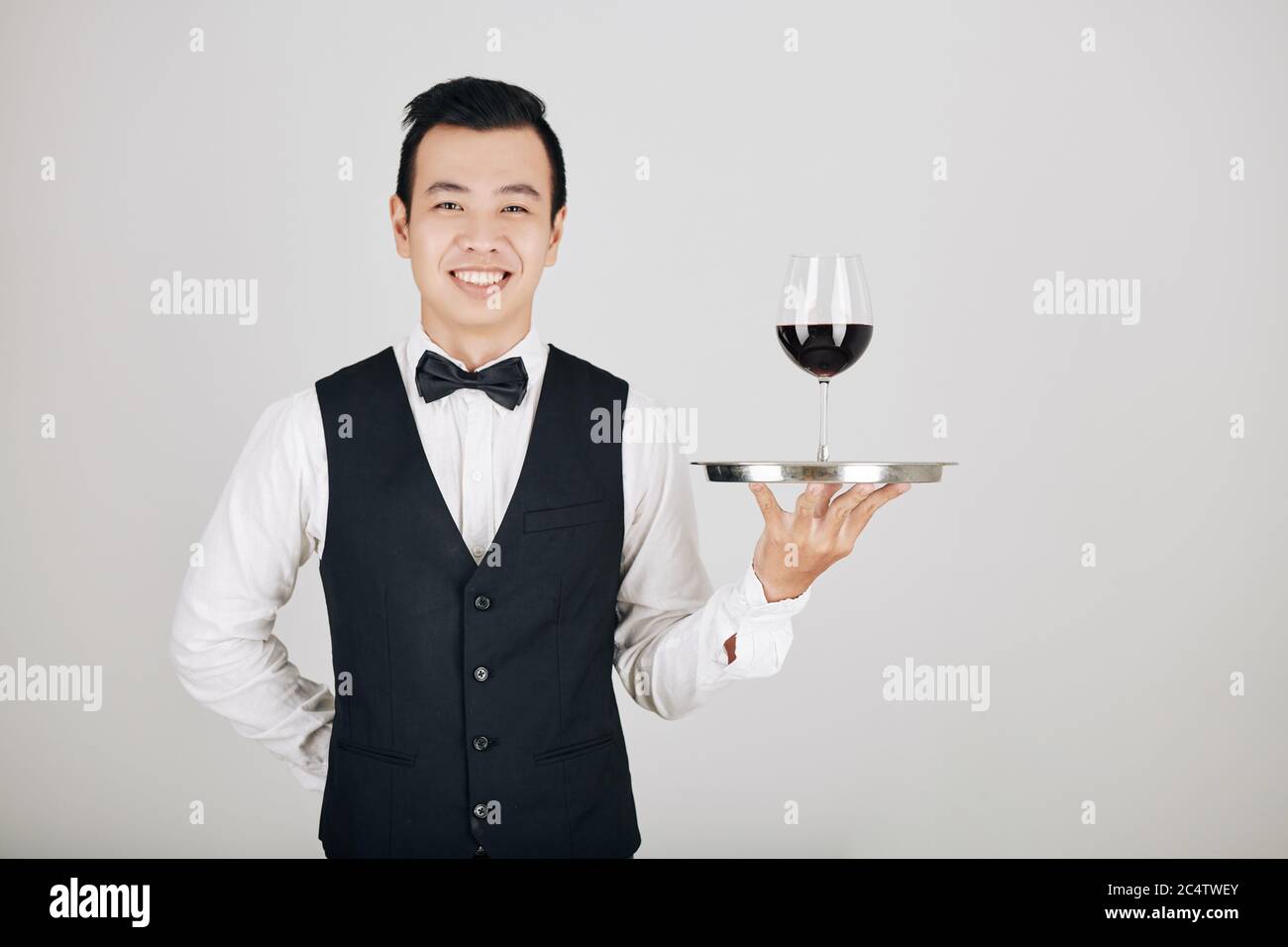 Portrait of smiling young Asian waiter holding tray with glass of red ...