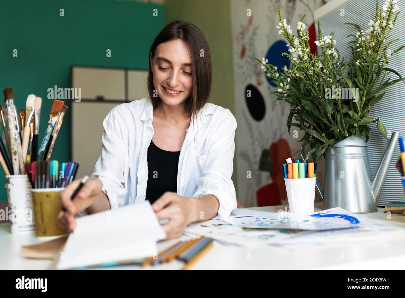 Smiling girl with dark hair sitting at the desk with paintings while ...
