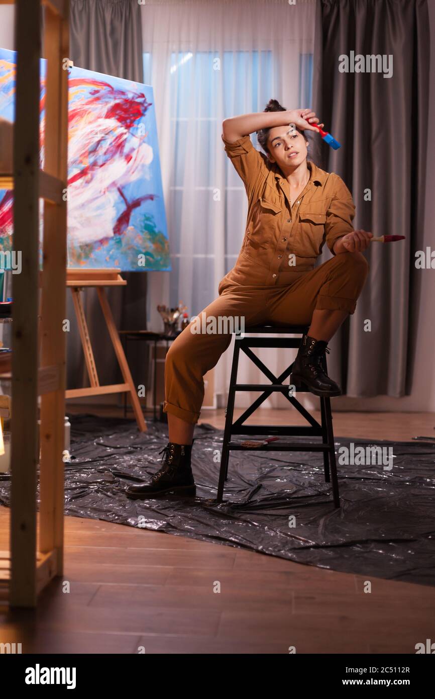 Overworked artist in studio sitting on chair holding paint brush ...