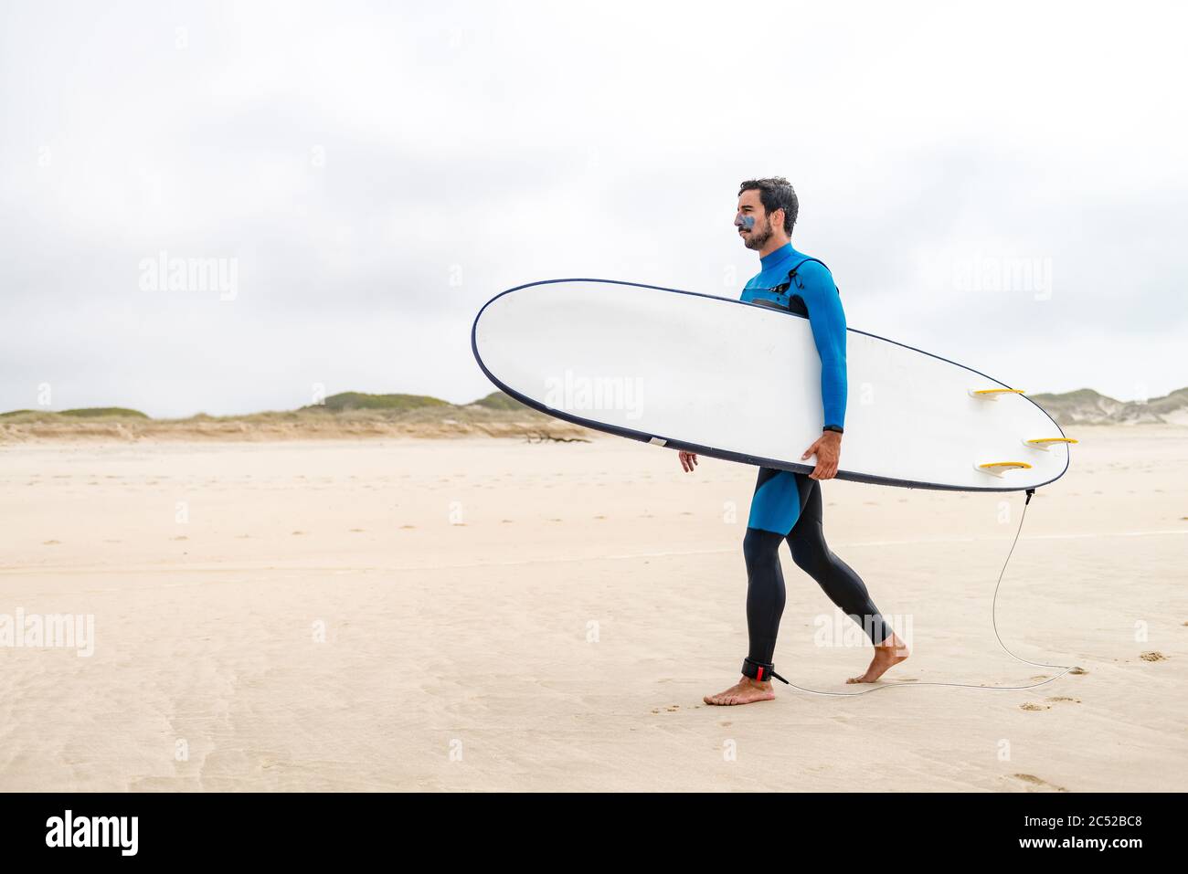 Young male surfer wearing wetsuit, holding surfboard under his arm ...