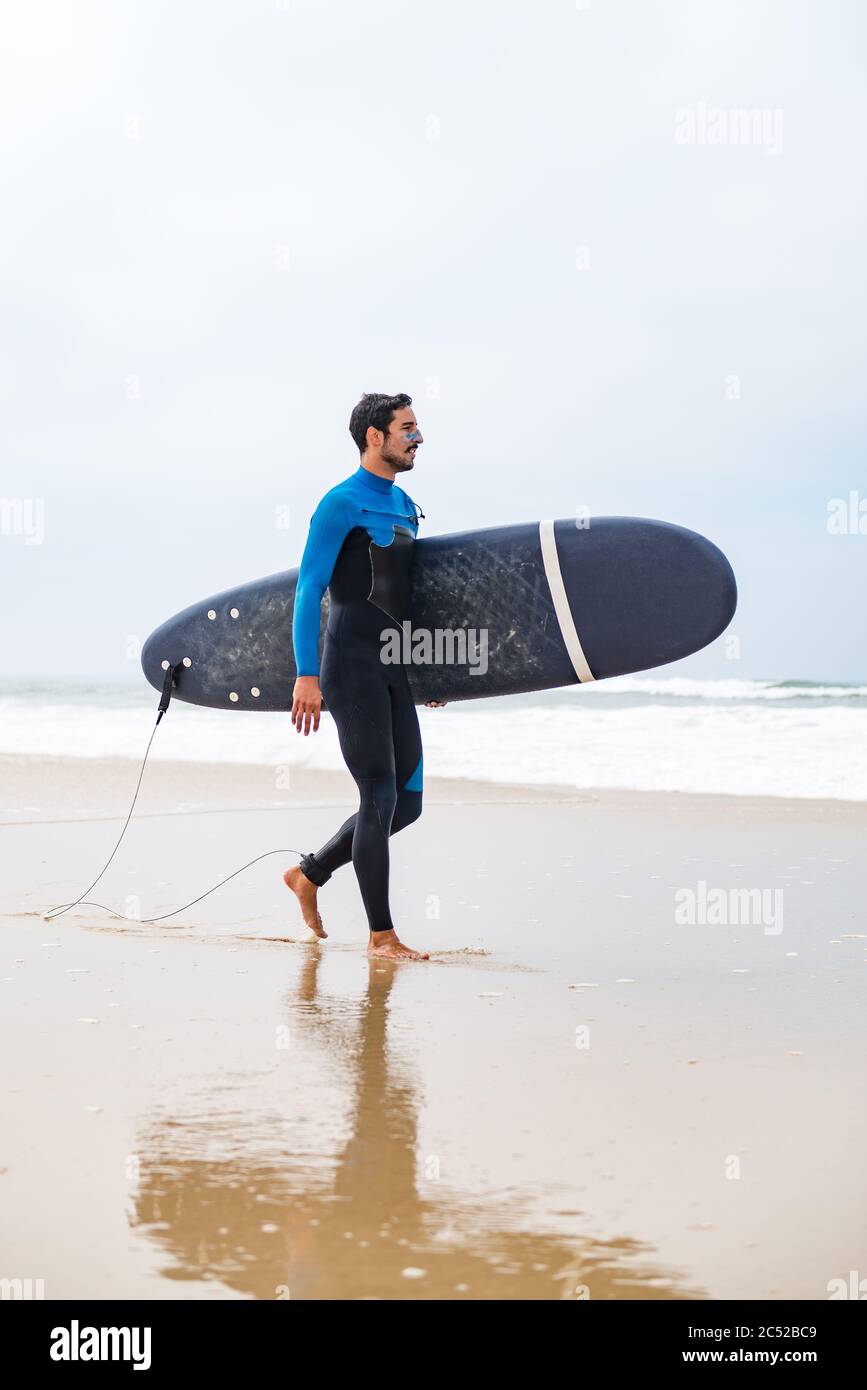 Young male surfer wearing wetsuit, holding surfboard under his arm ...