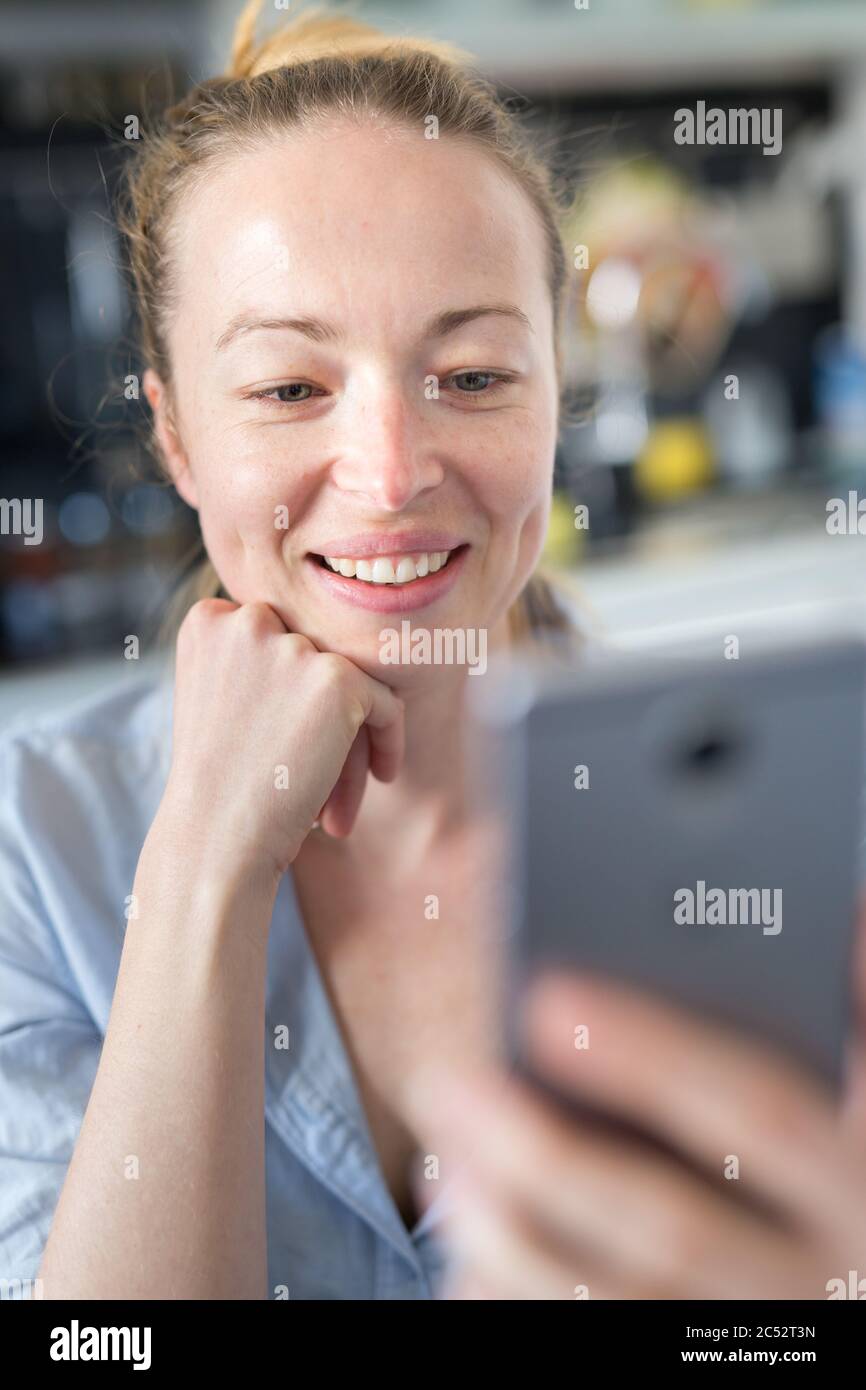 Young smiling cheerful pleased woman indoors at home kitchen using social media apps on mobile ...