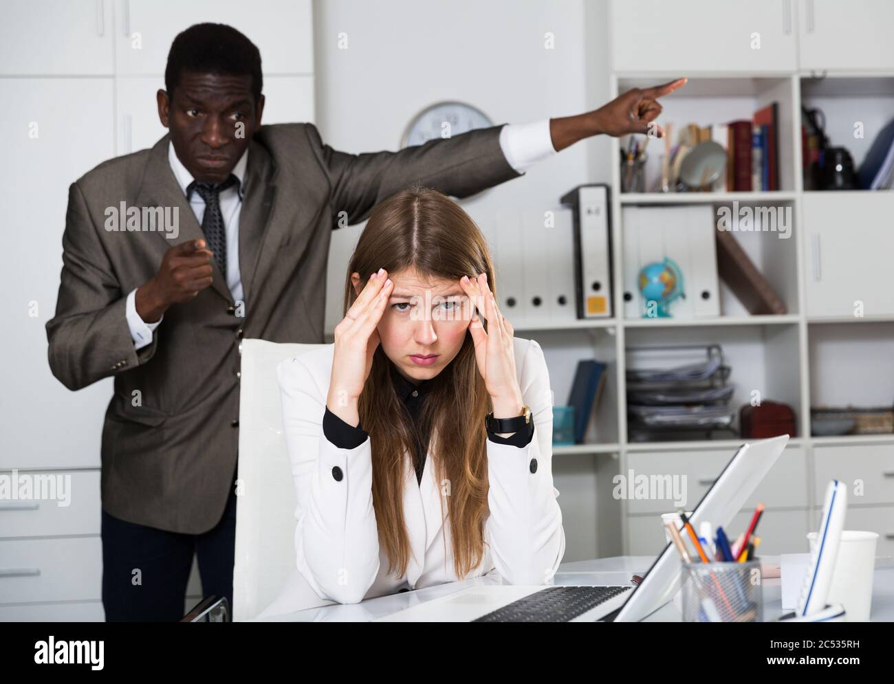 Frustrated woman sitting at office desk with disgruntled boss behind ...