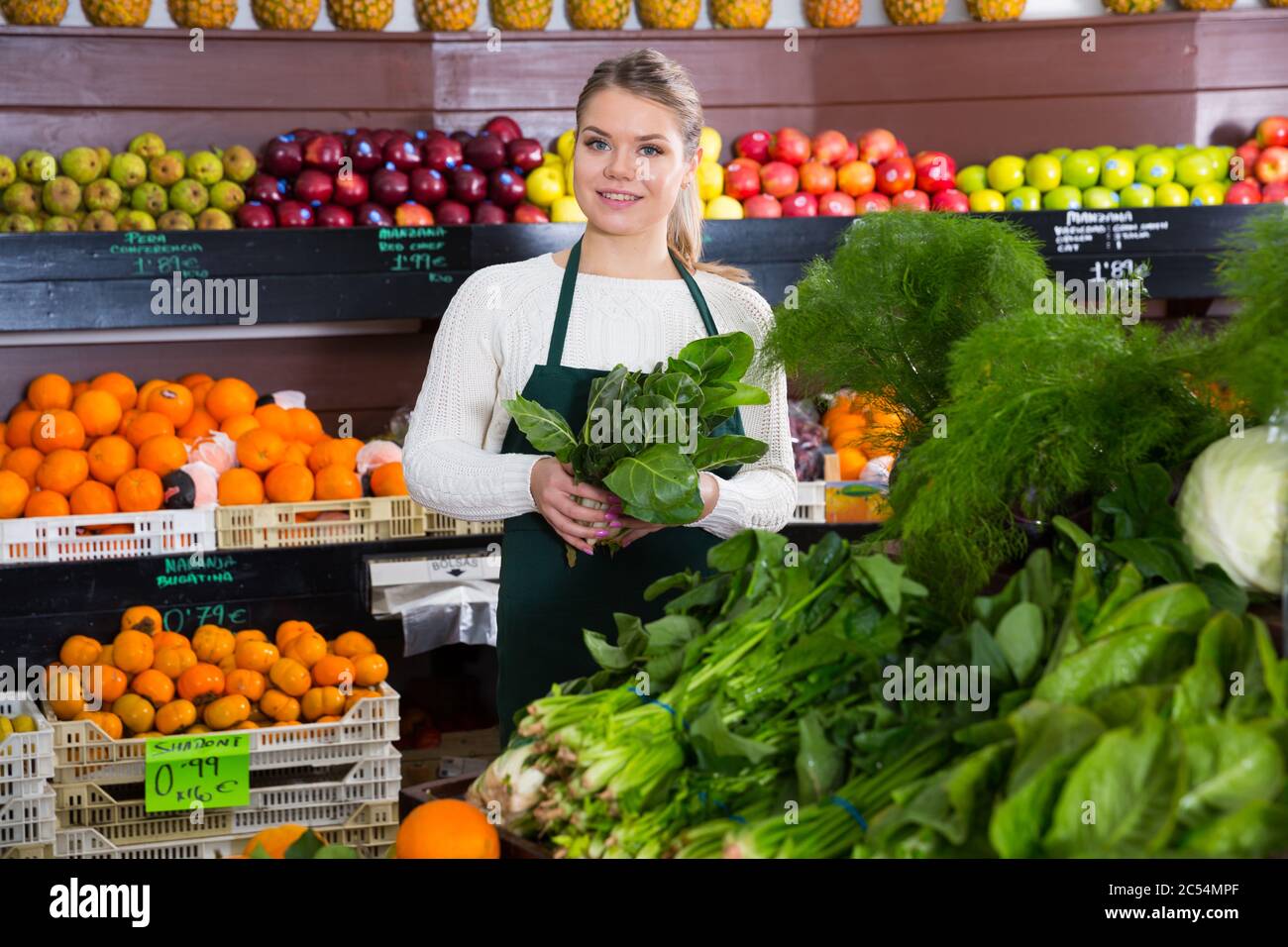 Positive female greengrocery worker in apron offering fresh greens in ...