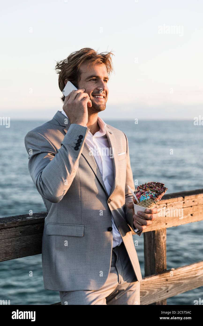 Businessman with ice cream cone talking on smart phone on ocean pier ...