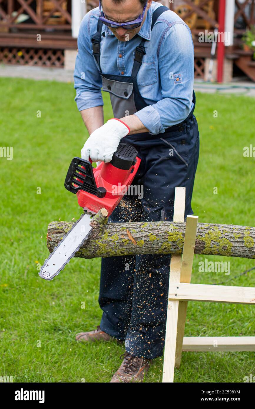 man in overalls saws wood with chain saw using sawhorse on green lawn ...