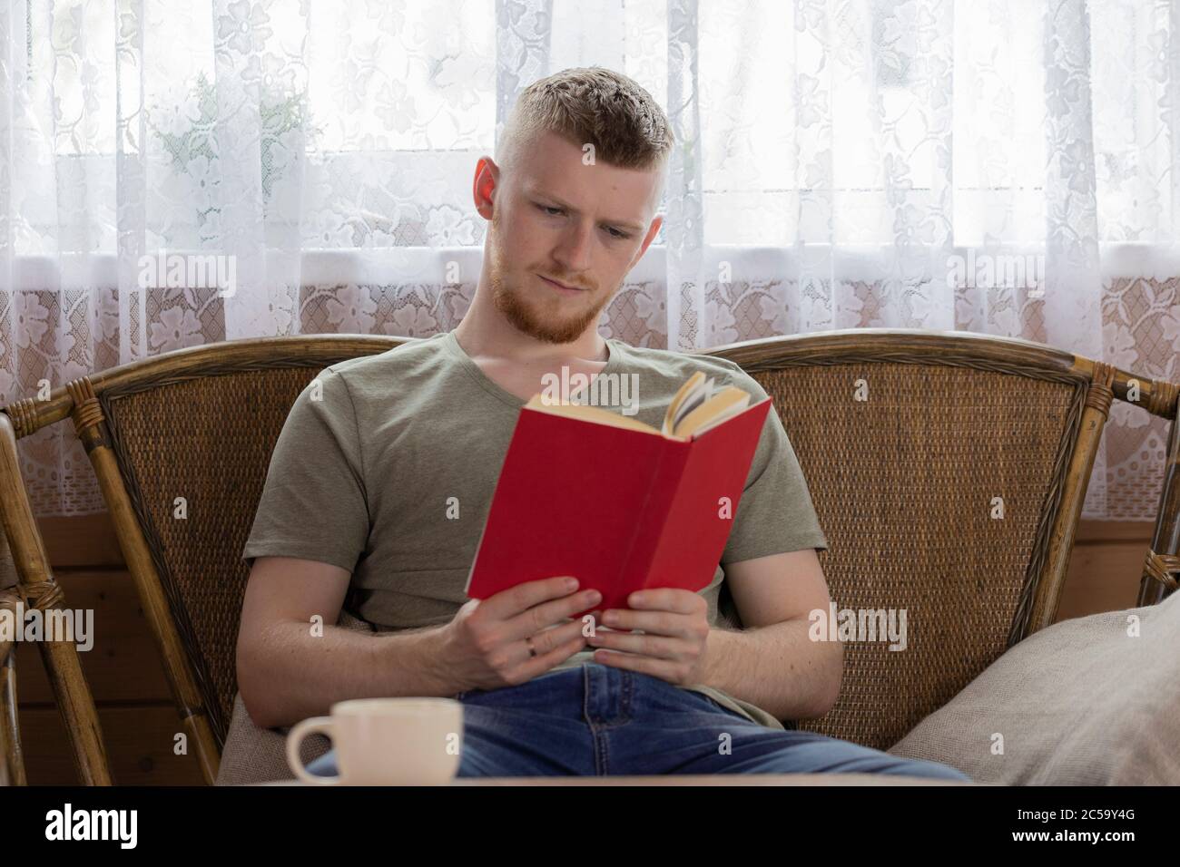 young man reading book with red cover on wicker bench in wooden house ...