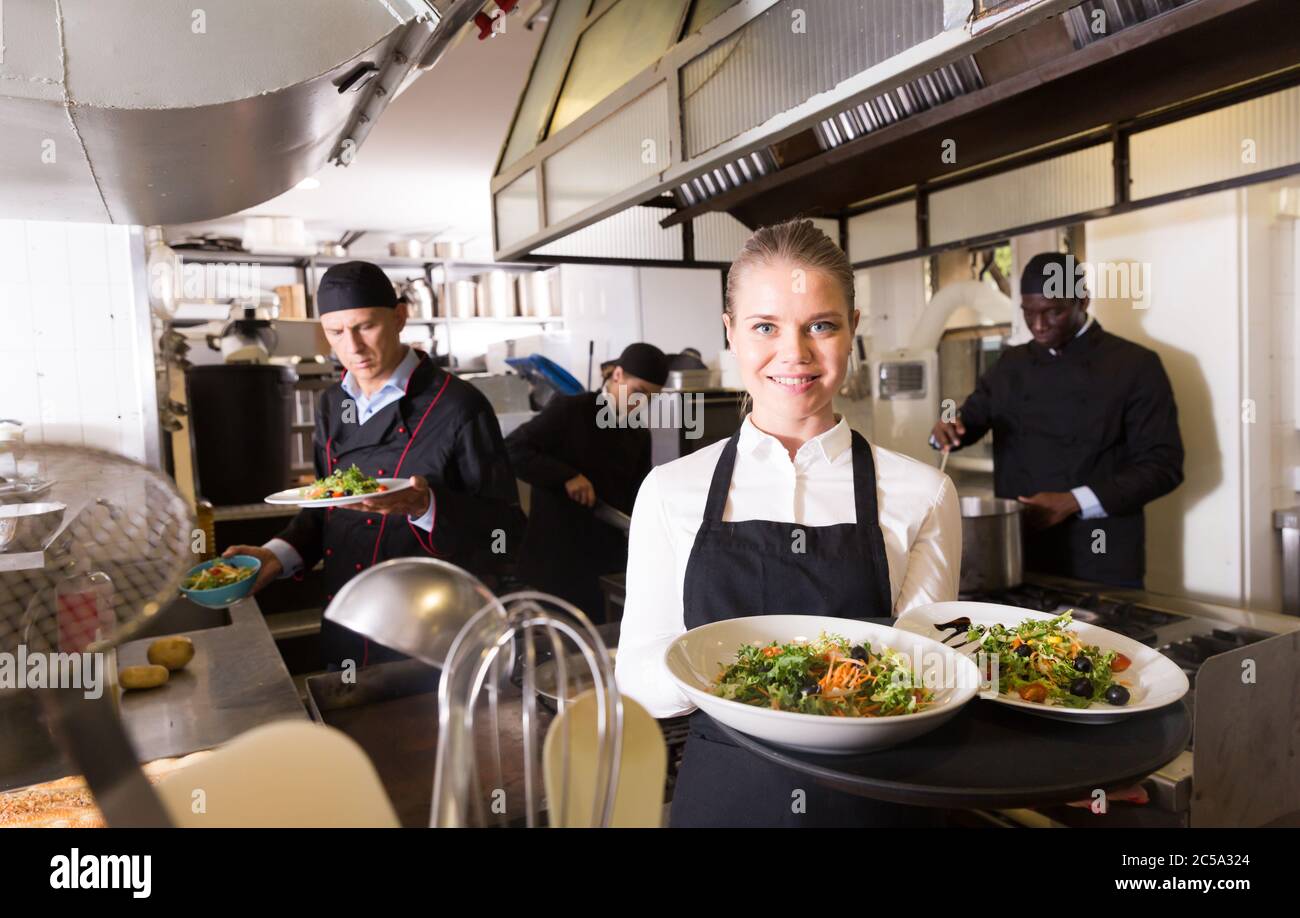 Portrait of attractive waitress with ordered dishes in kitchen of ...