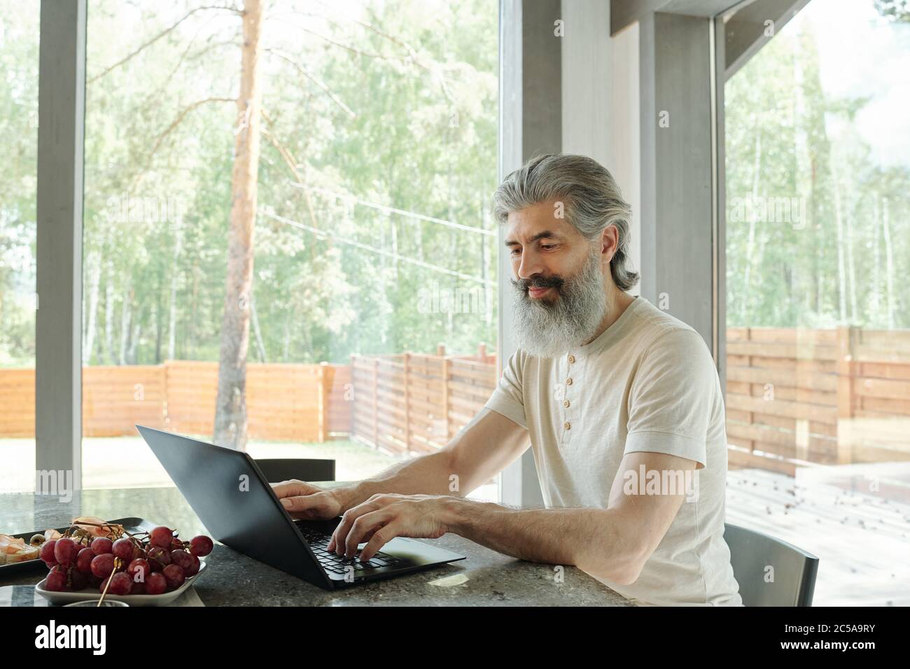 Content modern gray-bearded man sitting at kitchen counter and using ...