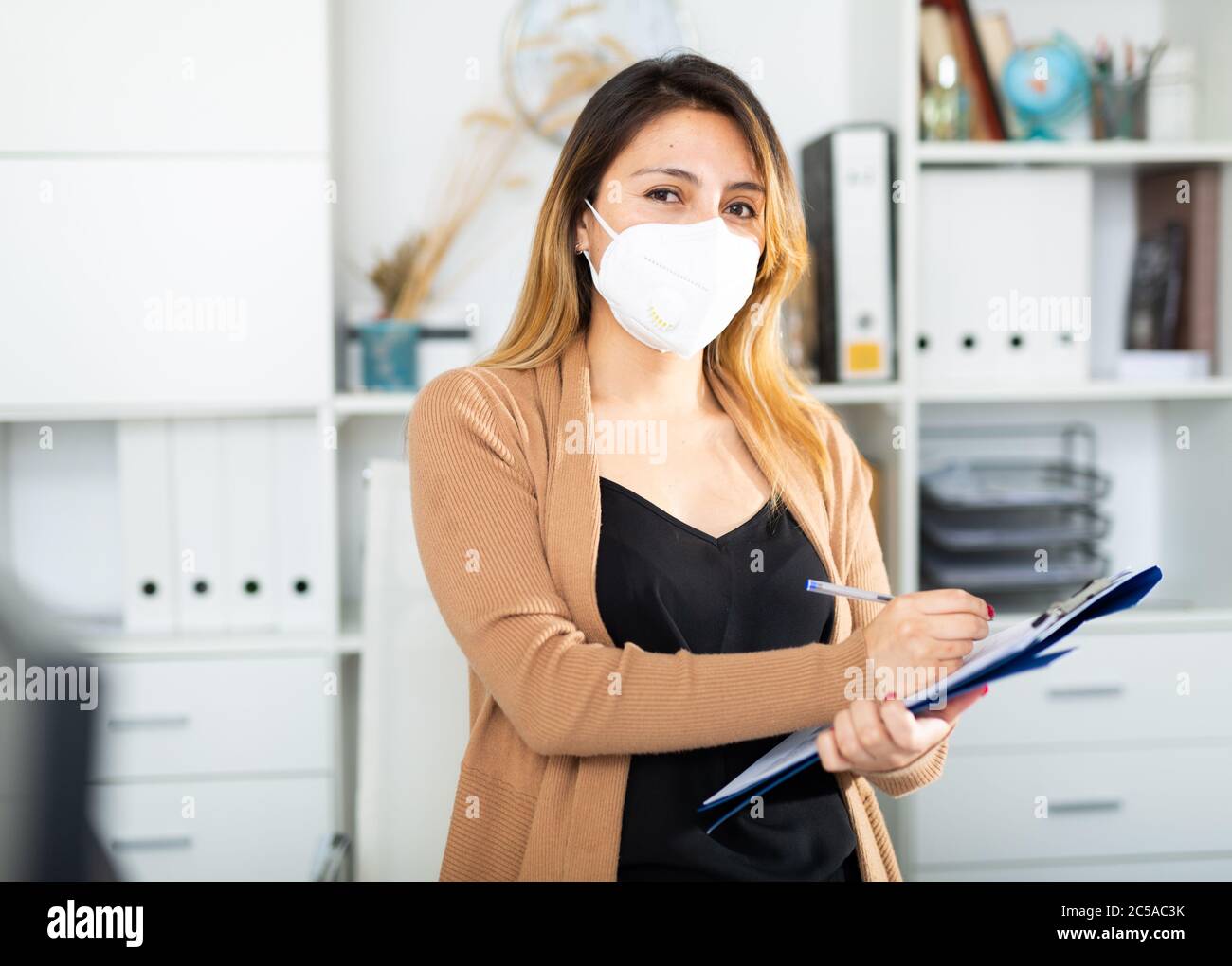 Young latina woman secretary wearing face mask standing in office with ...