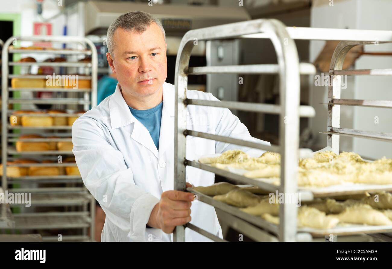 Experienced adult man working in bakery, pushing rack trolley with ...