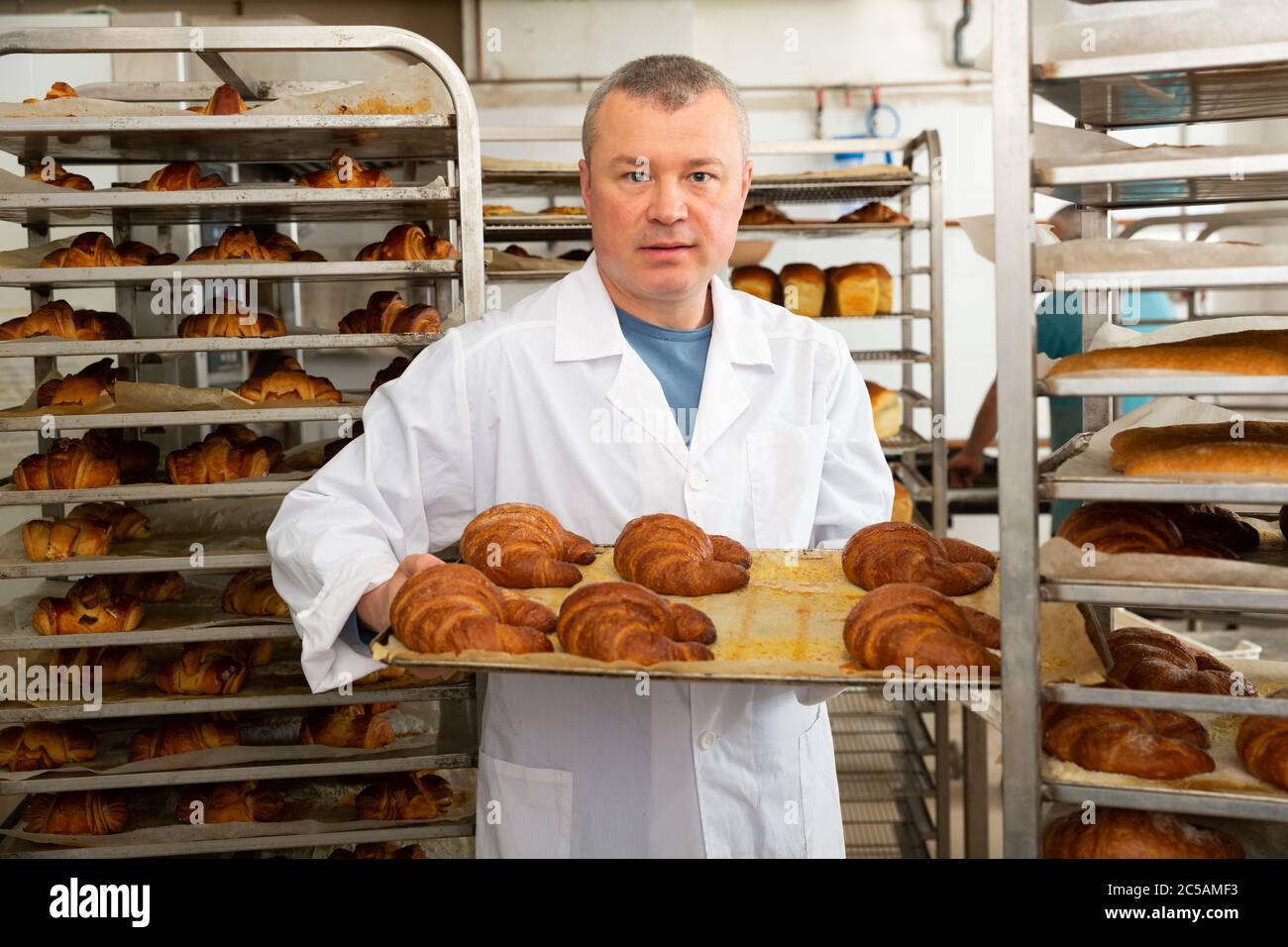 Portrait of positive baker working in bakehouse, putting tray with ...