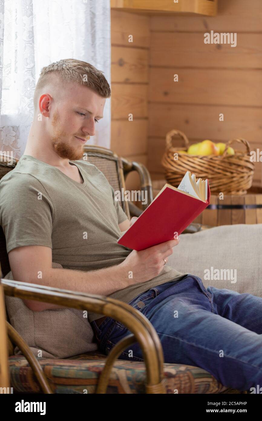 young man reading book with red cover on wicker bench in countryside ...