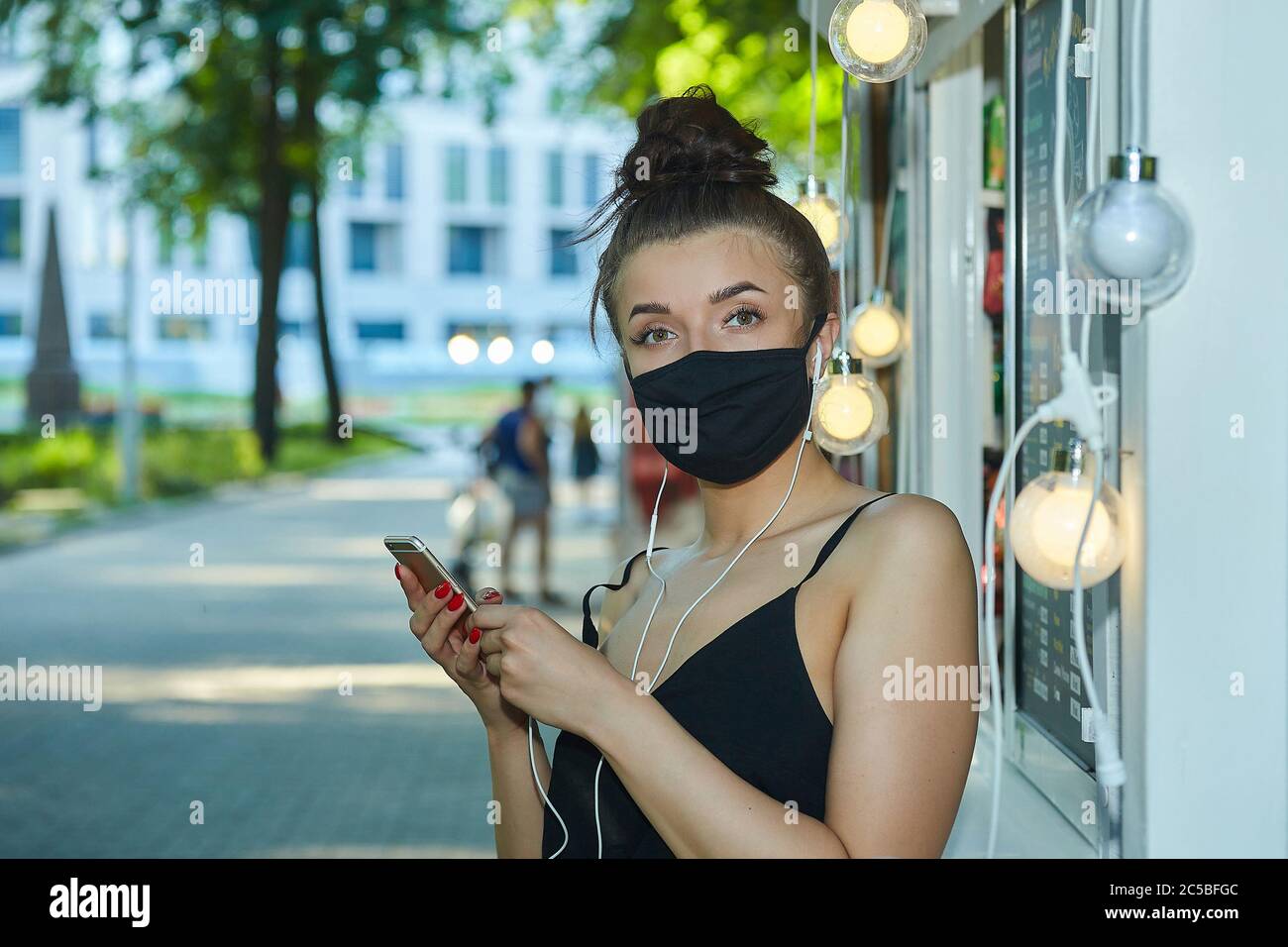 beautiful girl in a mask, posing in the city, holding a smartphone and ...