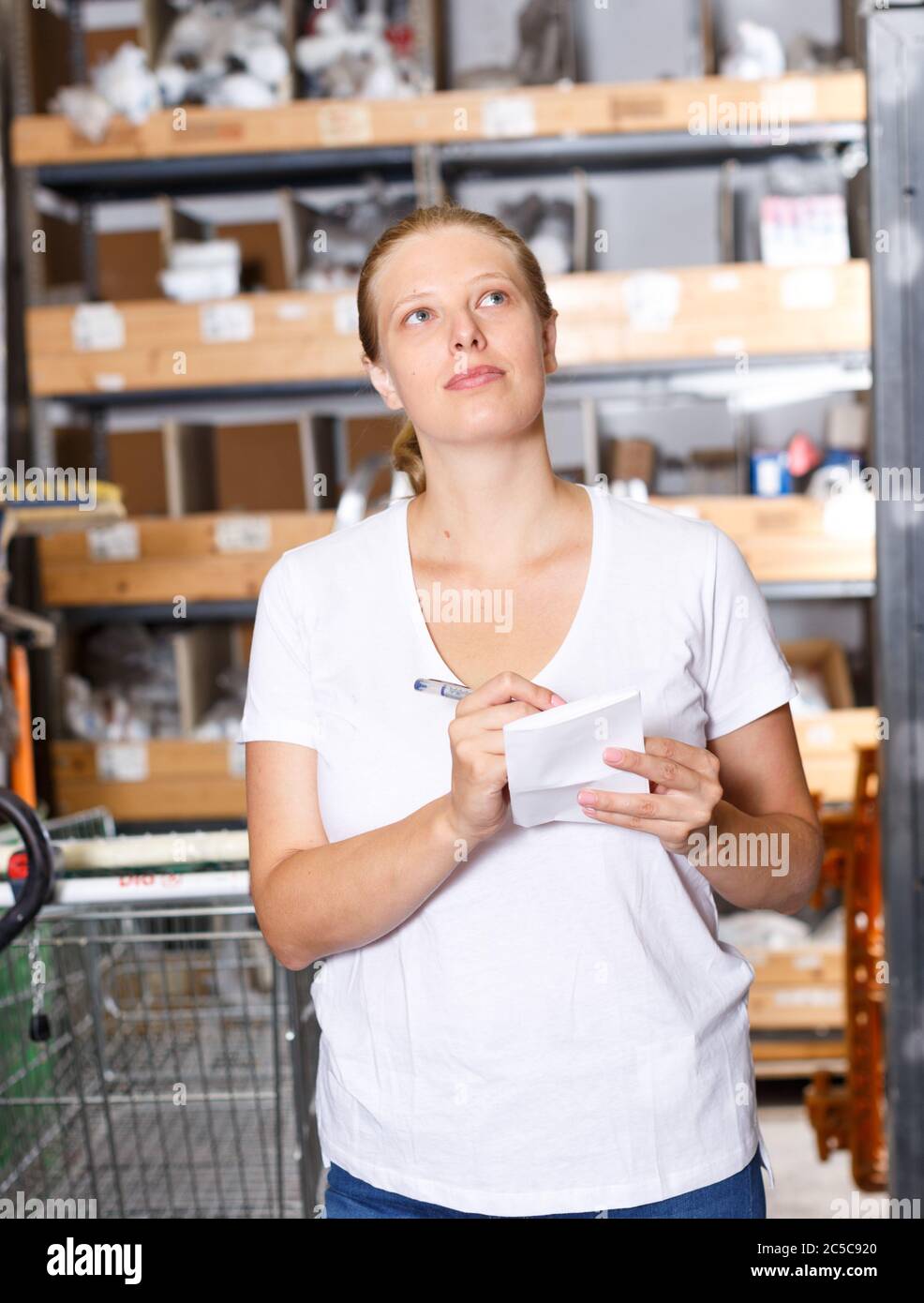 Young female customer holding shop list and looking tools at shelves in ...
