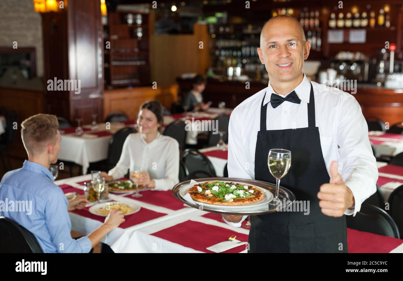 Polite waiter holding tray in restaurant with customers behind him ...
