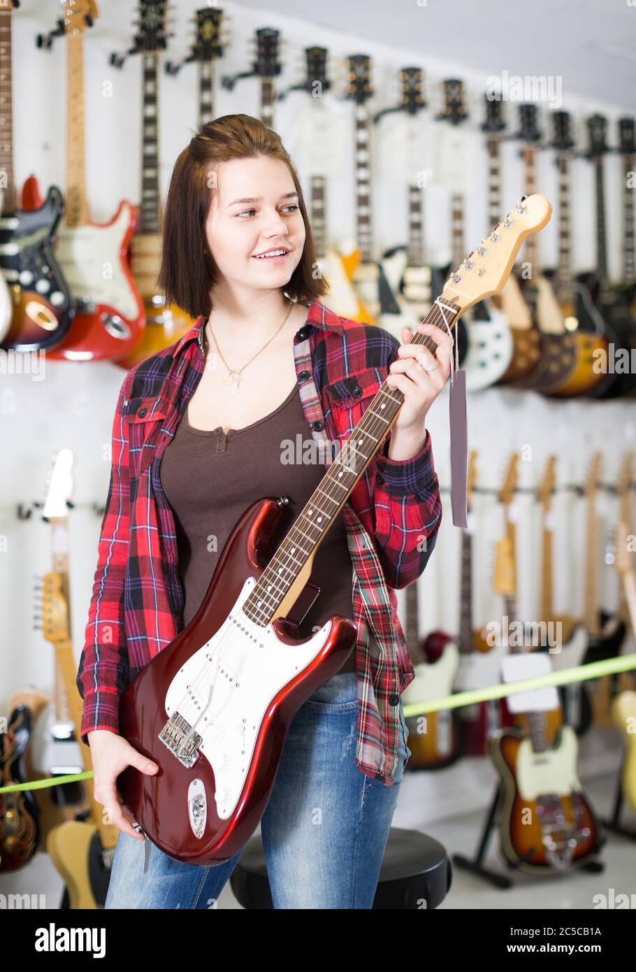 Сheerful teen girl examining various electric guitars in guitar shop ...