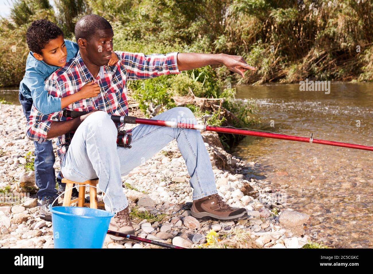Portrait of cheerful afro man and his little son fishing with rods on ...