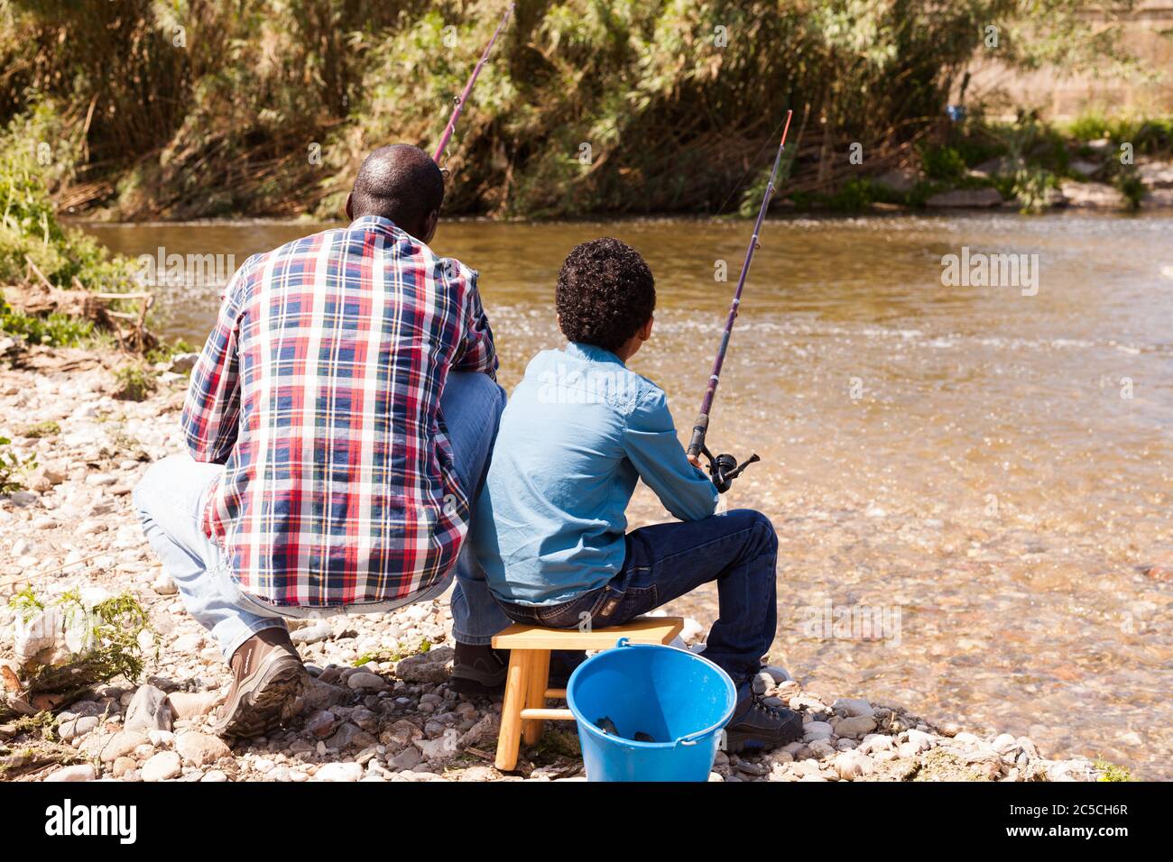 Back portrait of man and his little son sitting near river and fishing ...