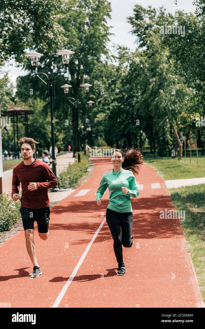 Handsome man running near positive girlfriend on running path in park ...
