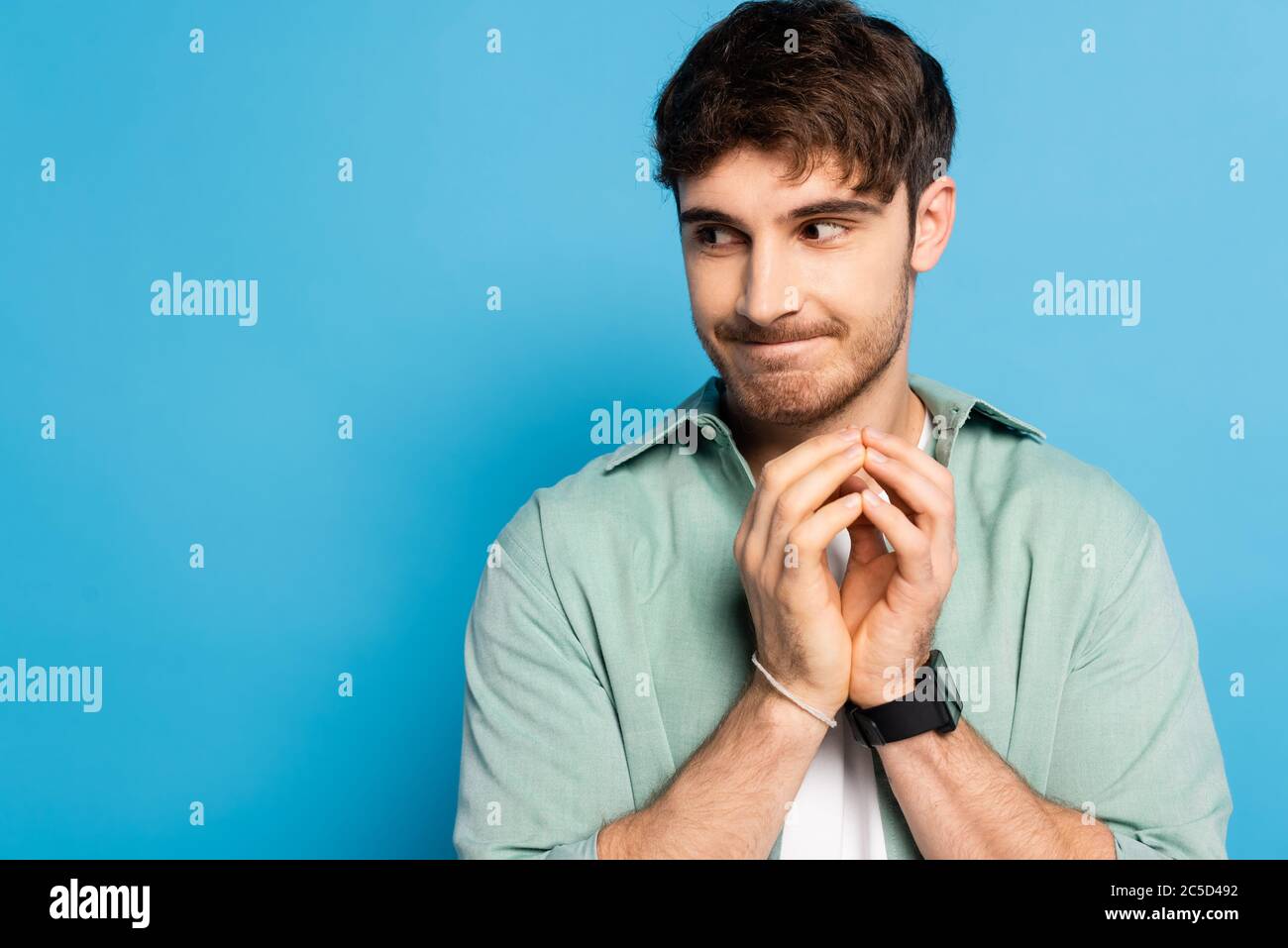 sly young man looking away while standing with joined fingers on blue ...