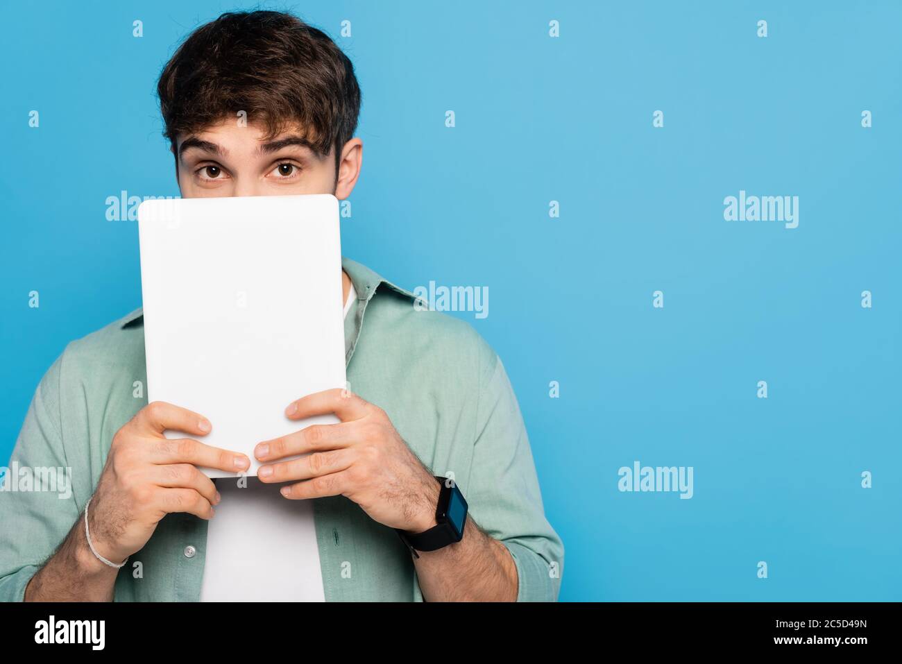scared young man obscuring face with digital tablet isolated on blue ...