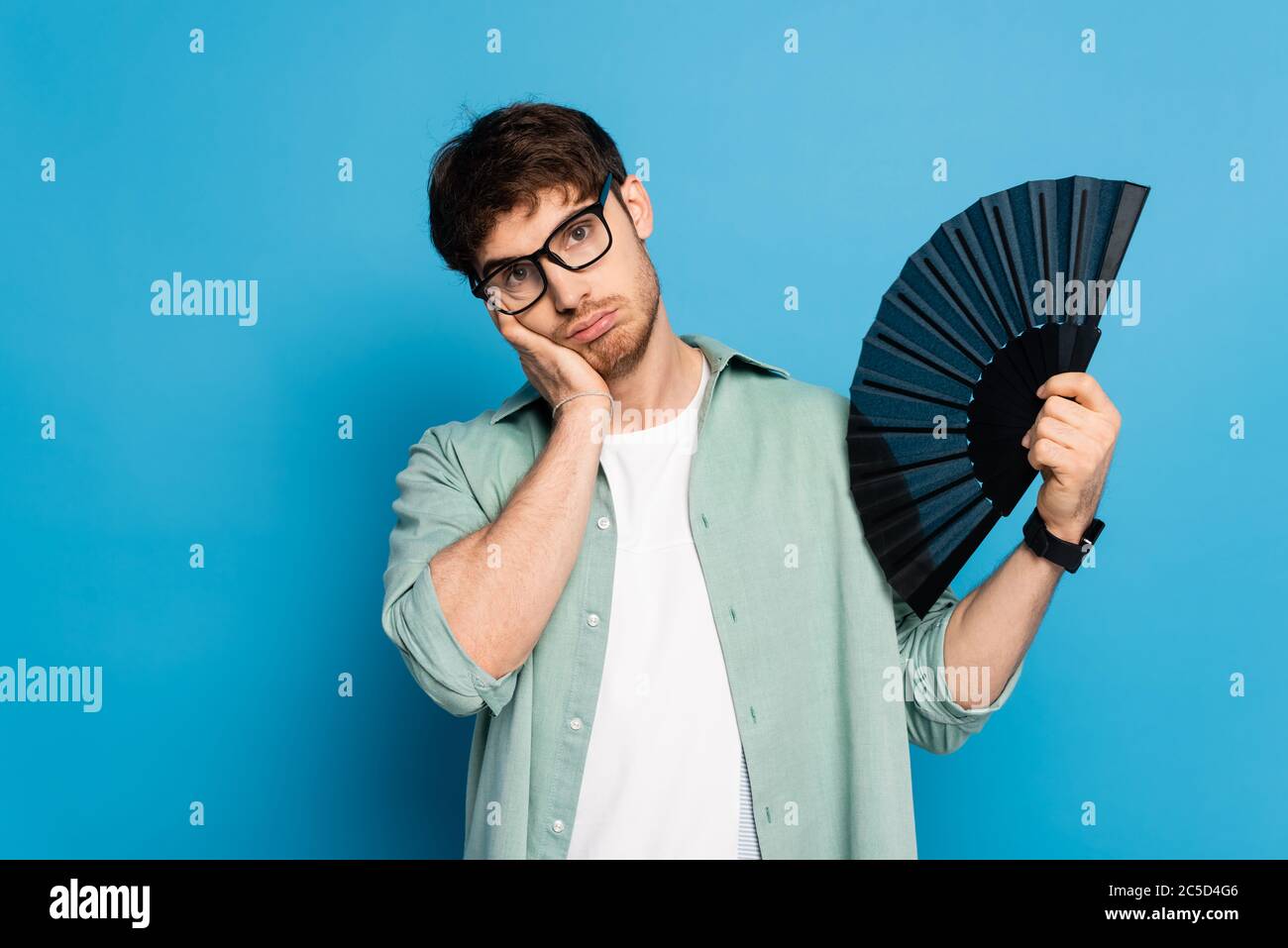 exhausted young man touching face while holding hand fan on blue Stock ...