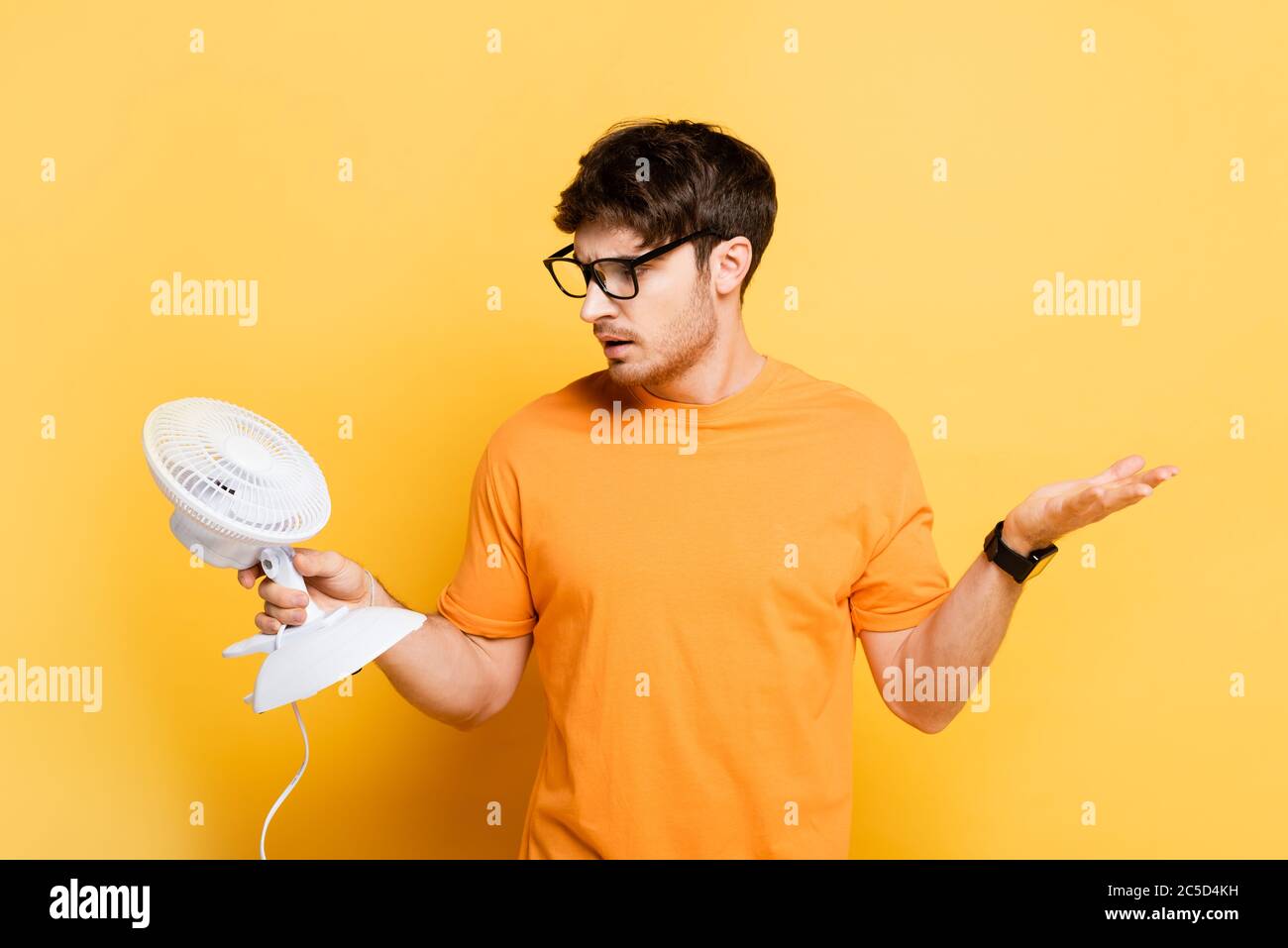 confused young man showing shrug gesture while holding electric fan on ...