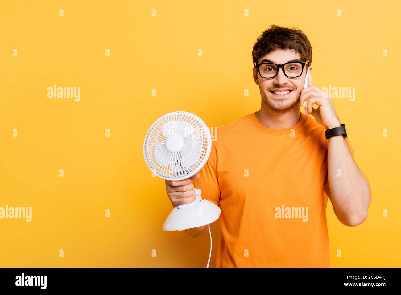 cheerful man talking on smartphone while holding electric fan on yellow ...