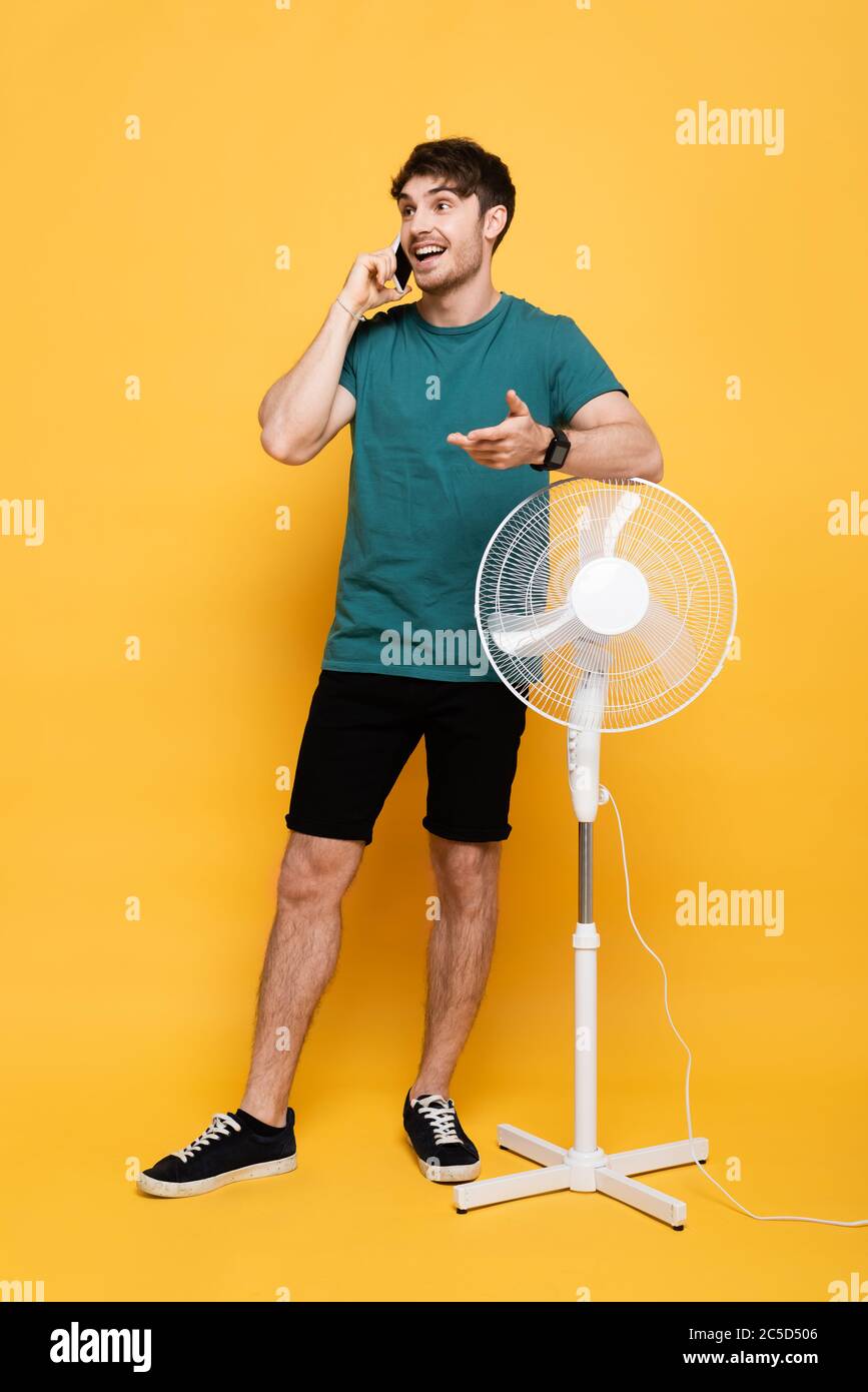 happy man talking on cellphone while standing near electric fan on ...