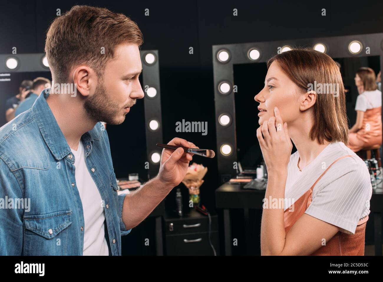 Side view of model pointing on cheek near makeup artist holding ...