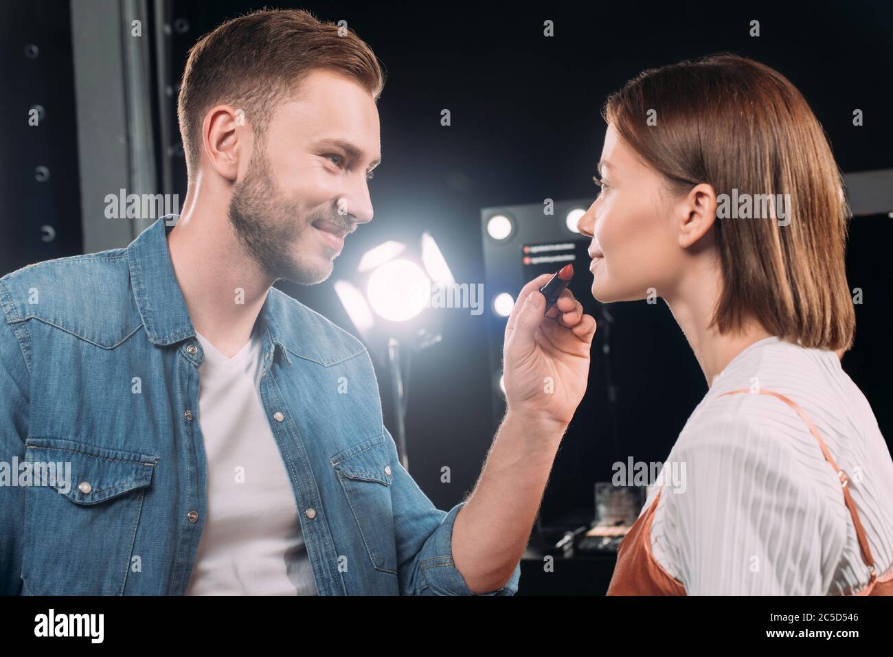 Handsome makeup artist smiling while applying lipstick on attractive ...
