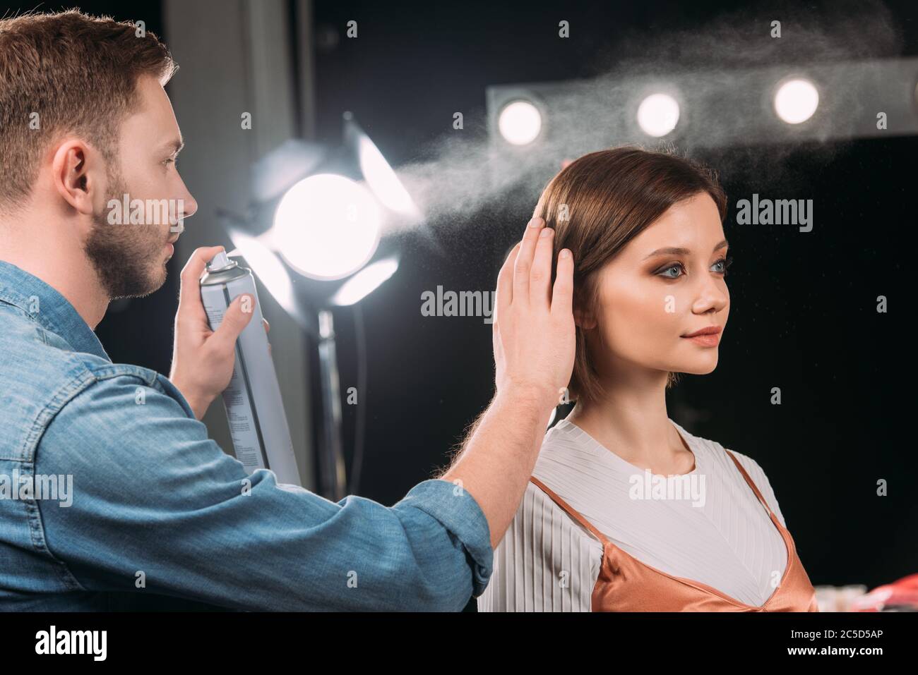 Handsome makeup artist using hair spray while doing hairstyle to ...