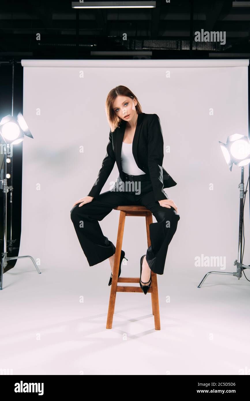 Elegant model in formal wear looking at camera while sitting on chair ...