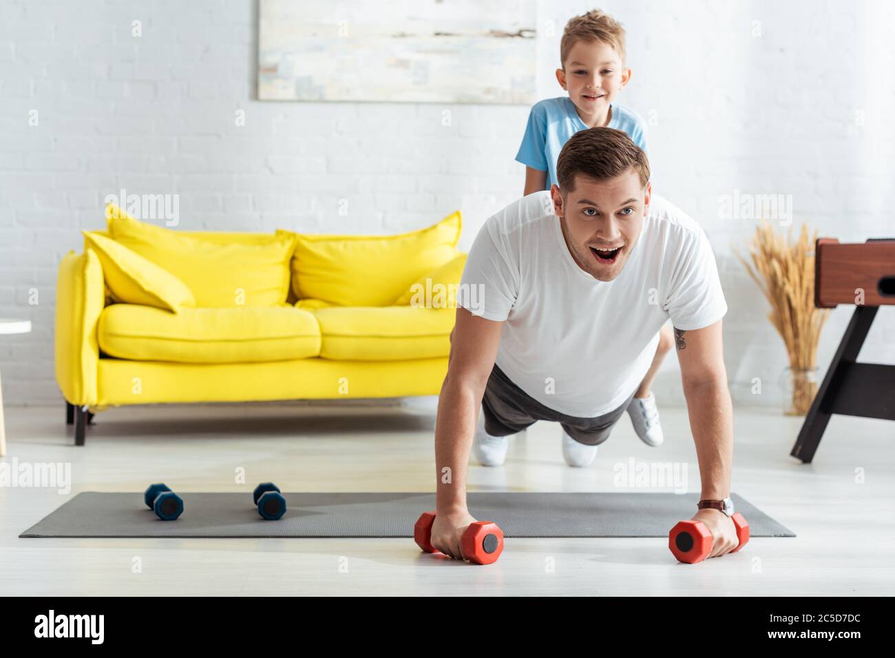 young man doing push ups with dumbbells while son sitting on his back ...