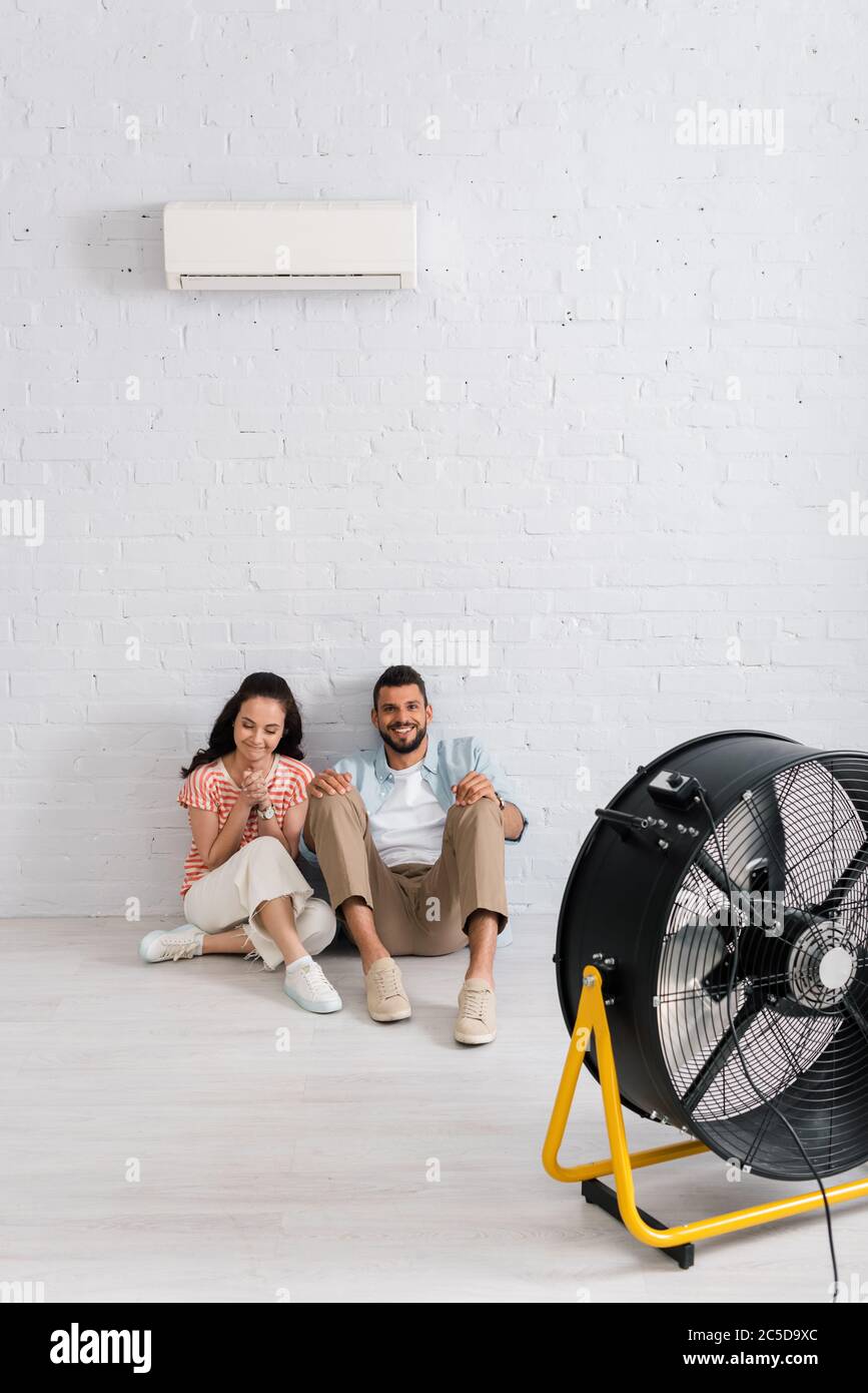Positive couple sitting on floor under air conditioner and electric fan ...