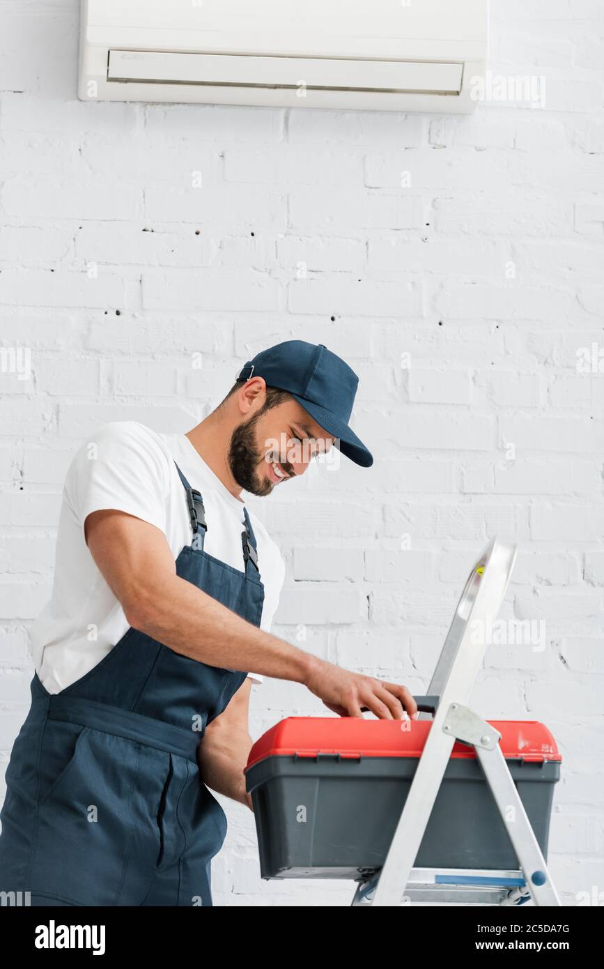 Smiling workman in uniform holding toolbox near ladder and air ...