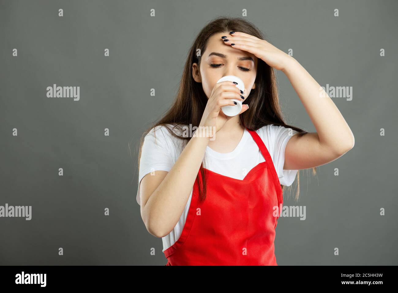 Portrait of young female supermarket employee drinking water on gray ...