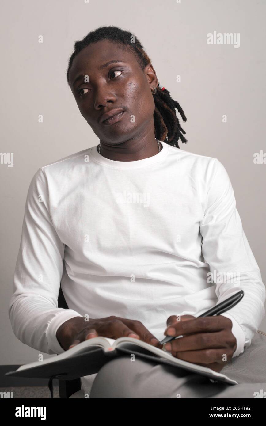 Young handsome African man with dreadlocks thinking while writing on ...