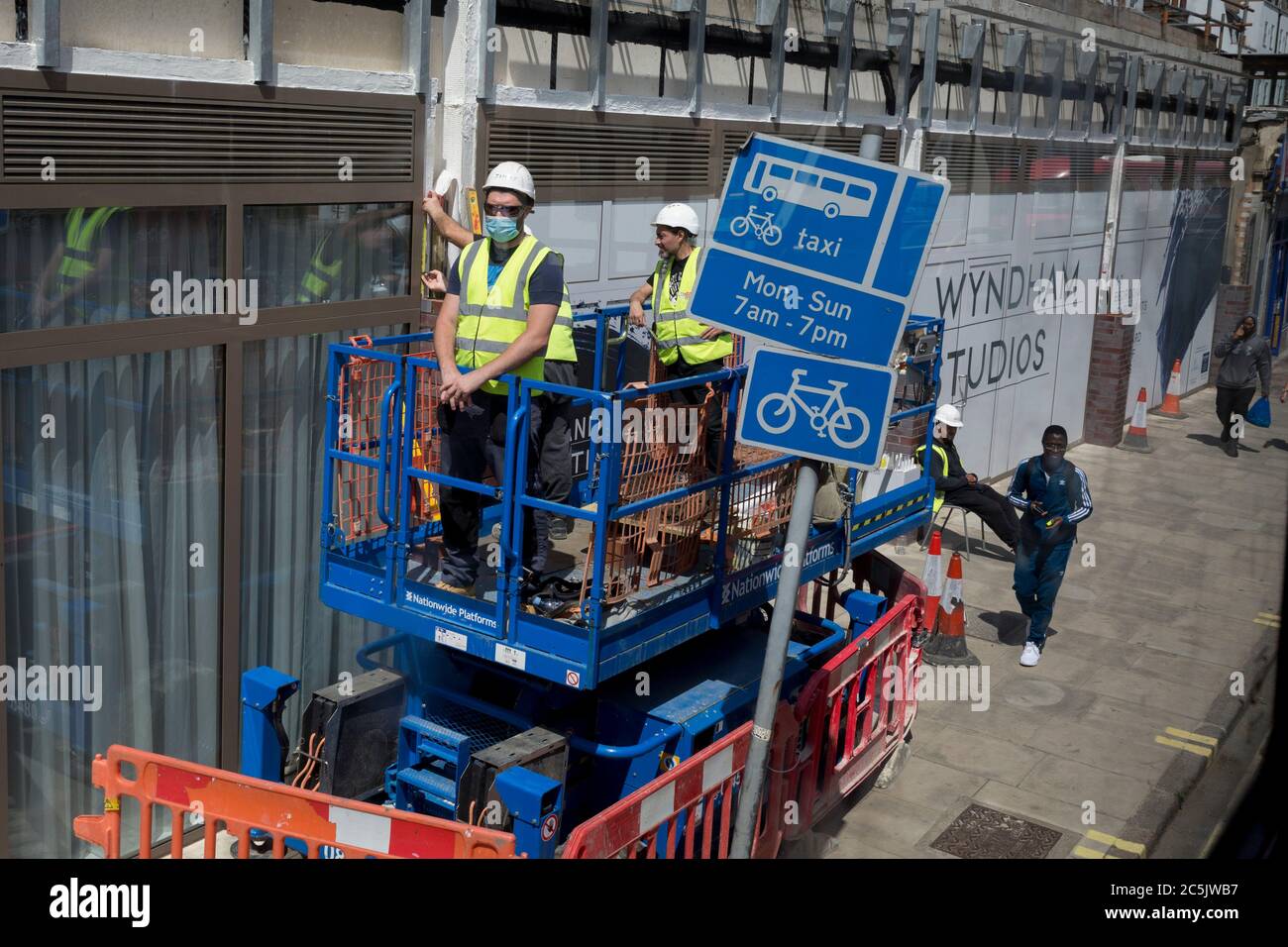 A workman wearing hi-visibility tabard and a face covering, stands as a ...